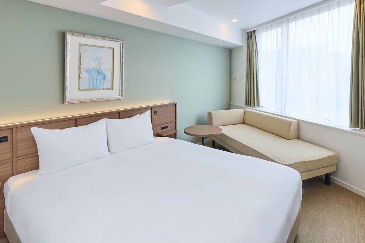 Quarto de hotel com uma cama grande, mesa de cabeceira, mesa redonda pequena, sofá e janela com cortinas.