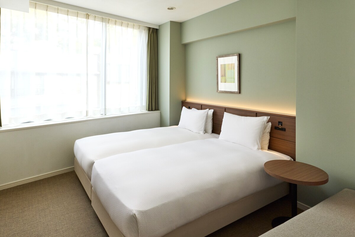 Quarto de hotel com duas camas, mesa de cabeceira, janela com cortinas e um quadro na parede.