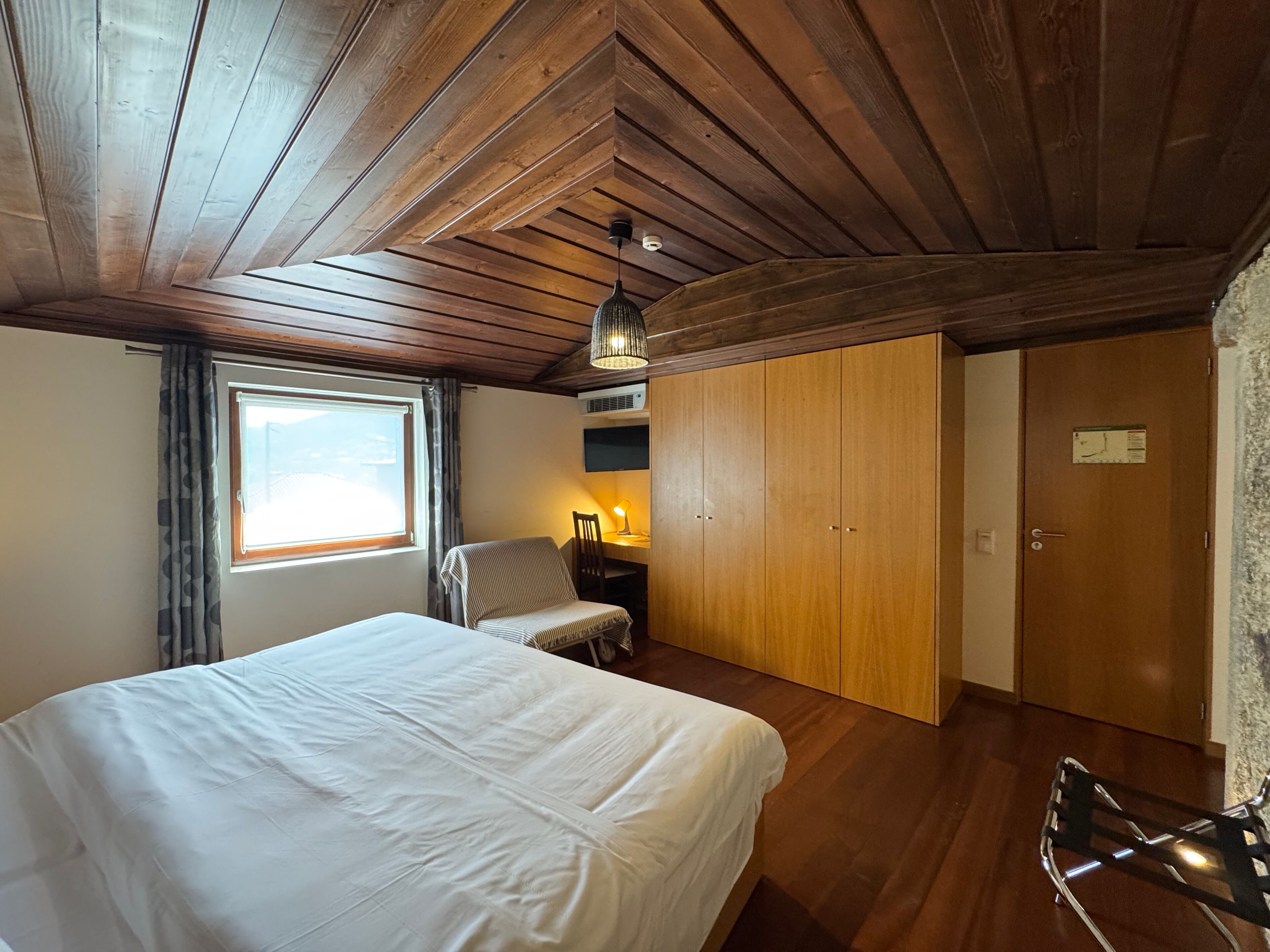 Quarto com teto de madeira, uma cama, uma cadeira, uma escrivaninha e um guarda-roupa.