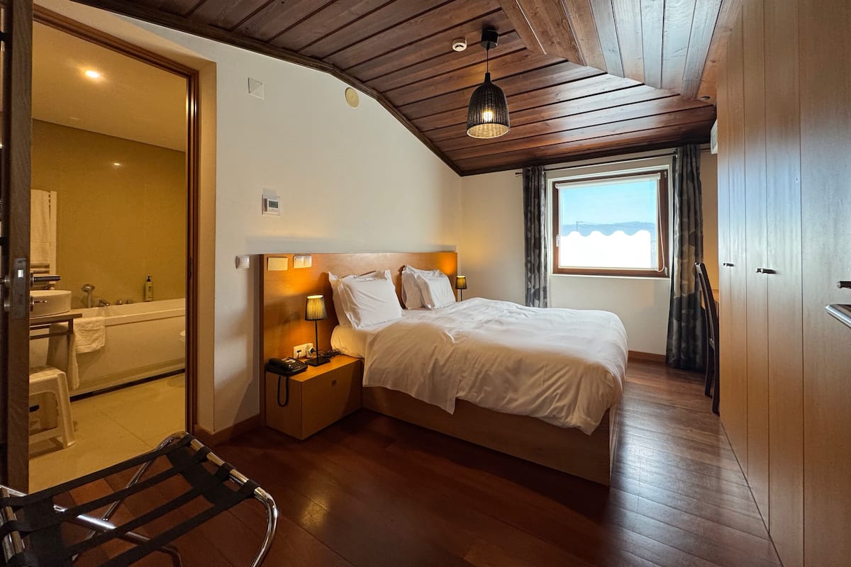 Quarto com teto de madeira, cama com roupa de cama branca, mesa de cabeceira com abajur, janela com vista e banheiro visível por uma porta aberta.