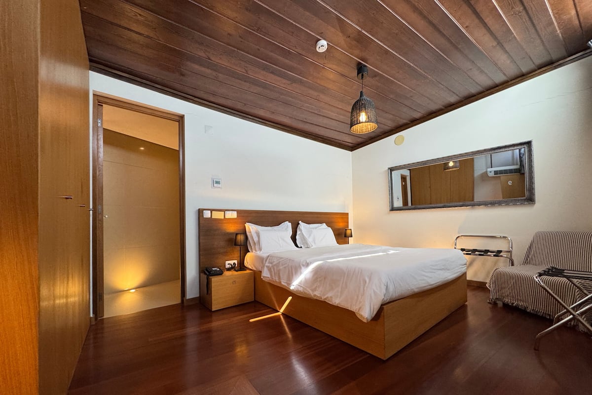 Quarto de hotel moderno com teto de madeira, uma cama grande com roupa de cama branca, uma mesinha de cabeceira com abajur, uma cadeira e um espelho na parede.
