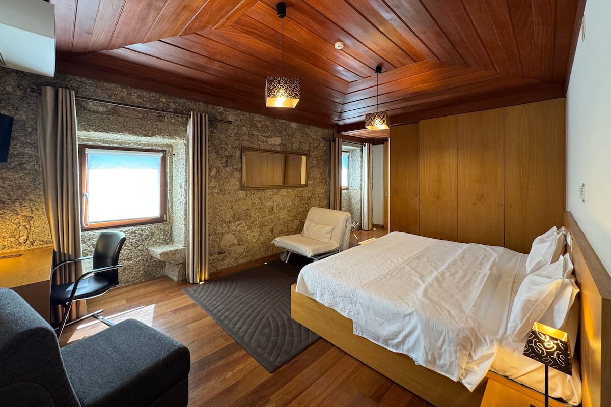 Quarto com teto de madeira, paredes de pedra, uma cama, uma cadeira e uma janela.