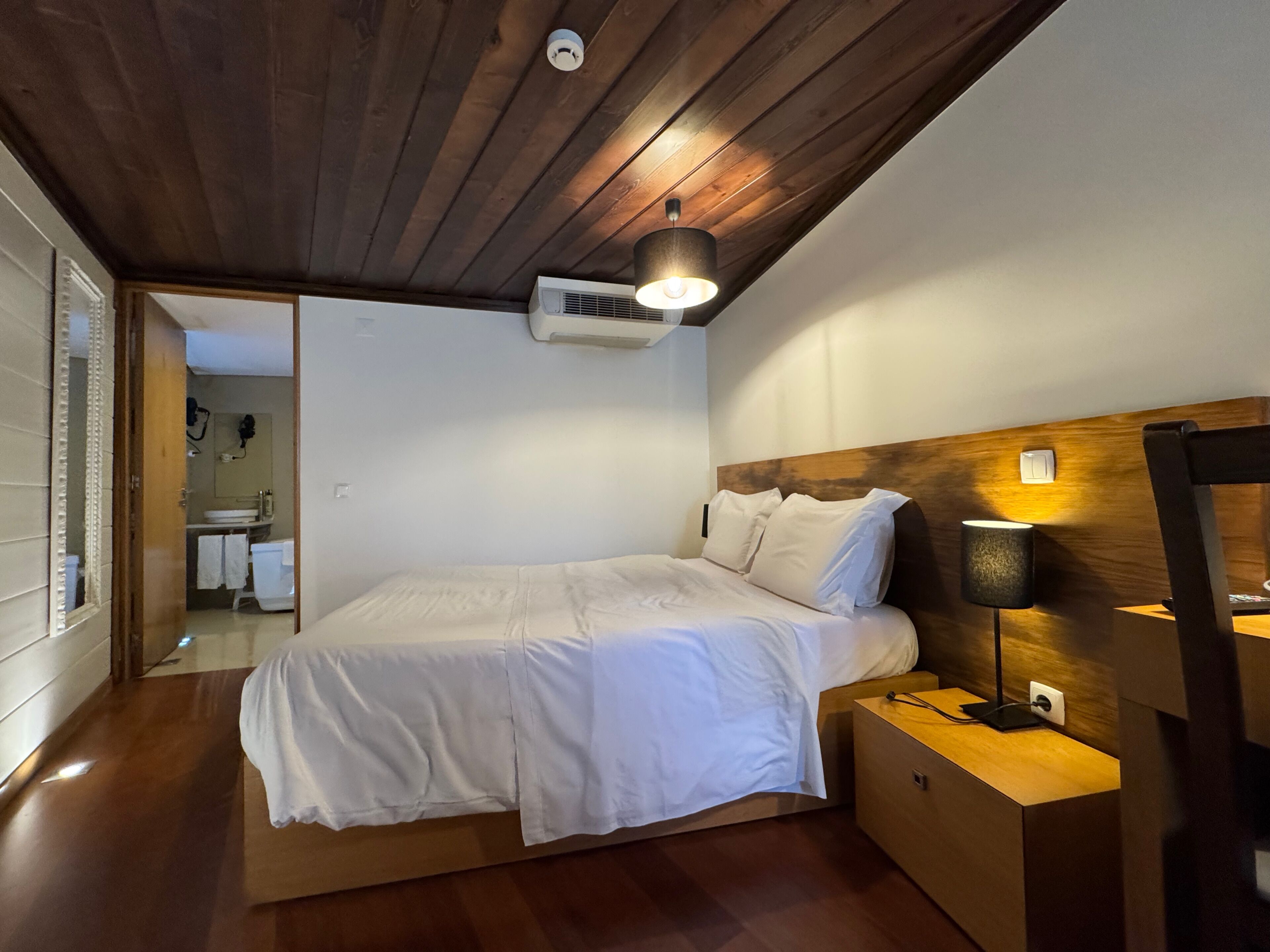 Quarto com teto de madeira, cama com roupa de cama branca, mesa de cabeceira com abajur e um pequeno armário de madeira.