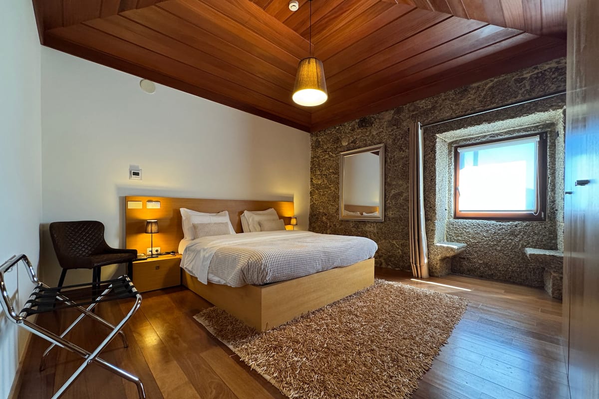 Quarto com cama, poltrona, mesa de cabeceira, luminária, parede de pedra e janela.