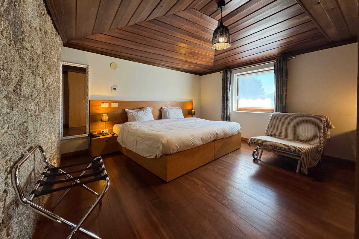 Quarto com teto de madeira, cama com lençóis brancos, uma cadeira, mesa de cabeceira e janela com cortinas.