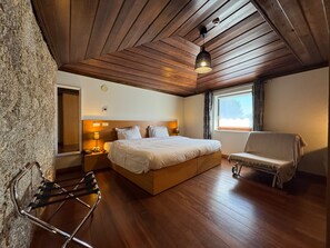 Quarto com teto de madeira, cama com lençóis brancos, uma cadeira, mesa de cabeceira e janela com cortinas.