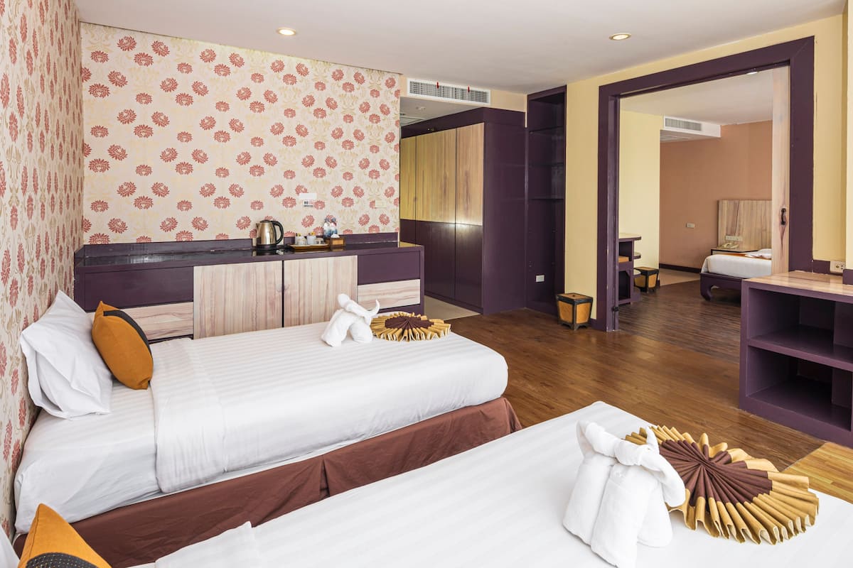 Quarto de hotel com duas camas, cabeceira de madeira, pequeno espaço com cozinha e banheiro visível por uma porta aberta.