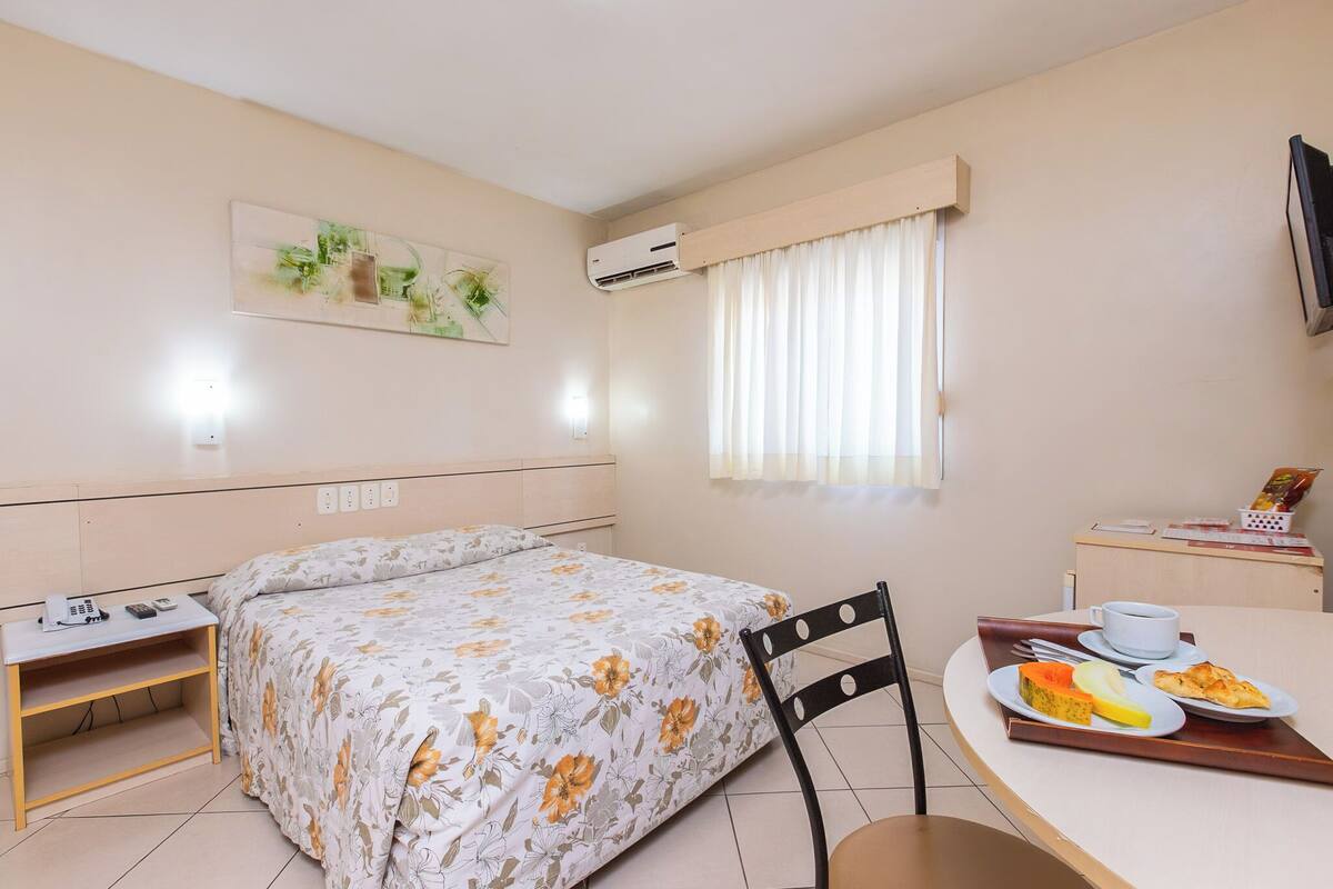 Quarto de hotel com cama, mesa de cabeceira, cadeira e bandeja de café da manhã com comida e uma xícara.