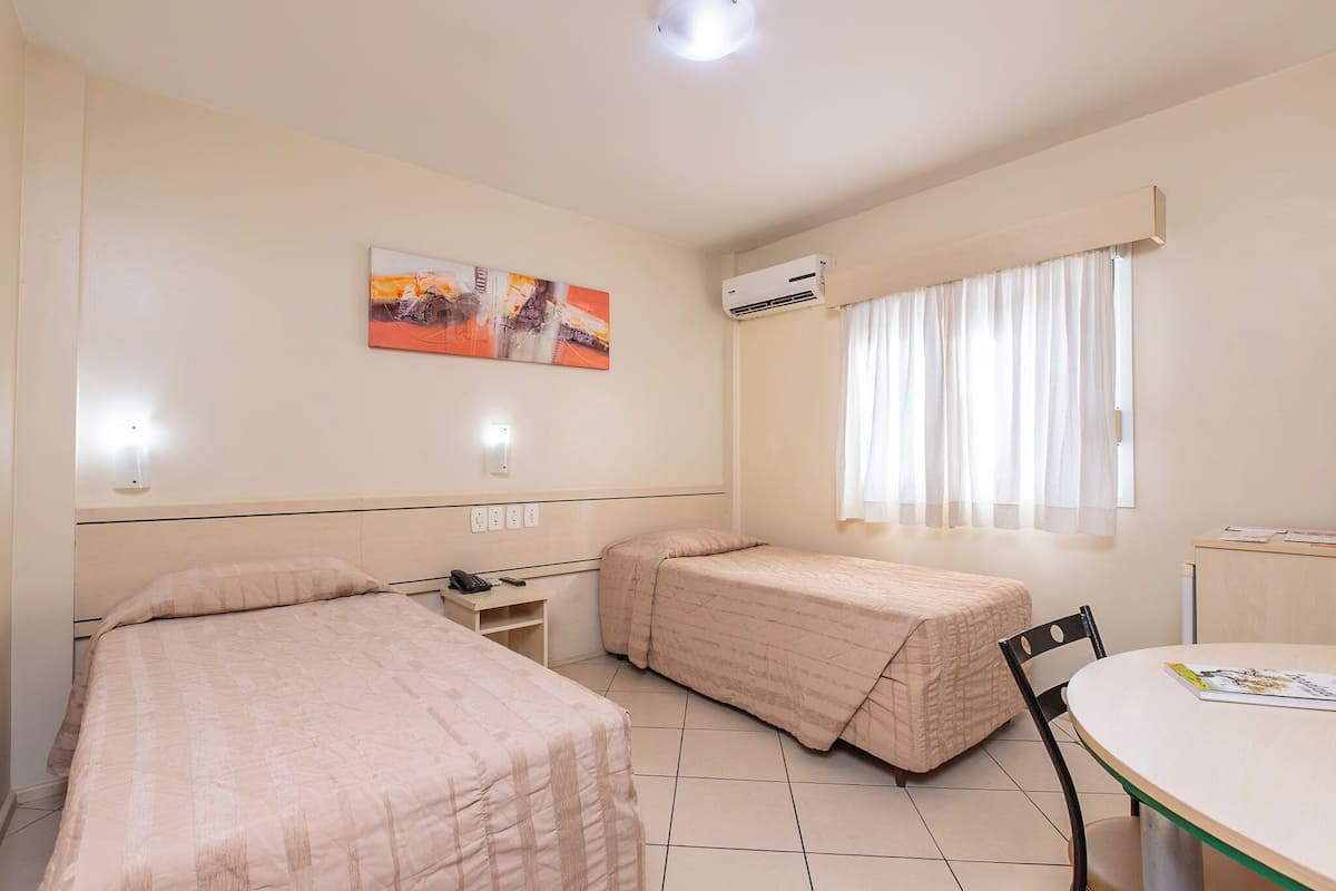 Quarto com duas camas, uma escrivaninha, uma cadeira, uma mesa, ar condicionado, janela com cortinas e um quadro na parede.