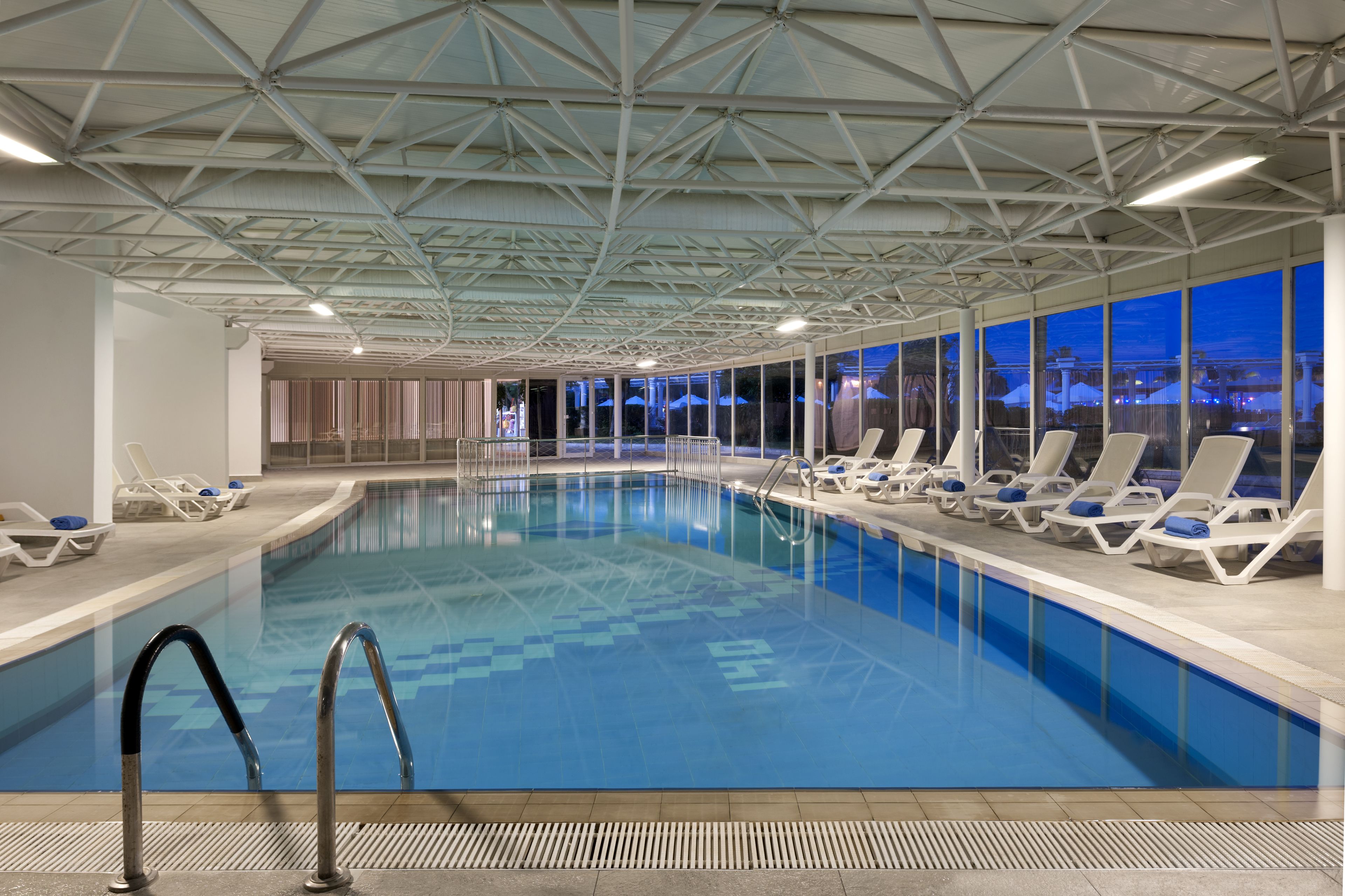 Piscina interna com água azul cristalina, rodeada por espreguiçadeiras e um teto alto com design geométrico.