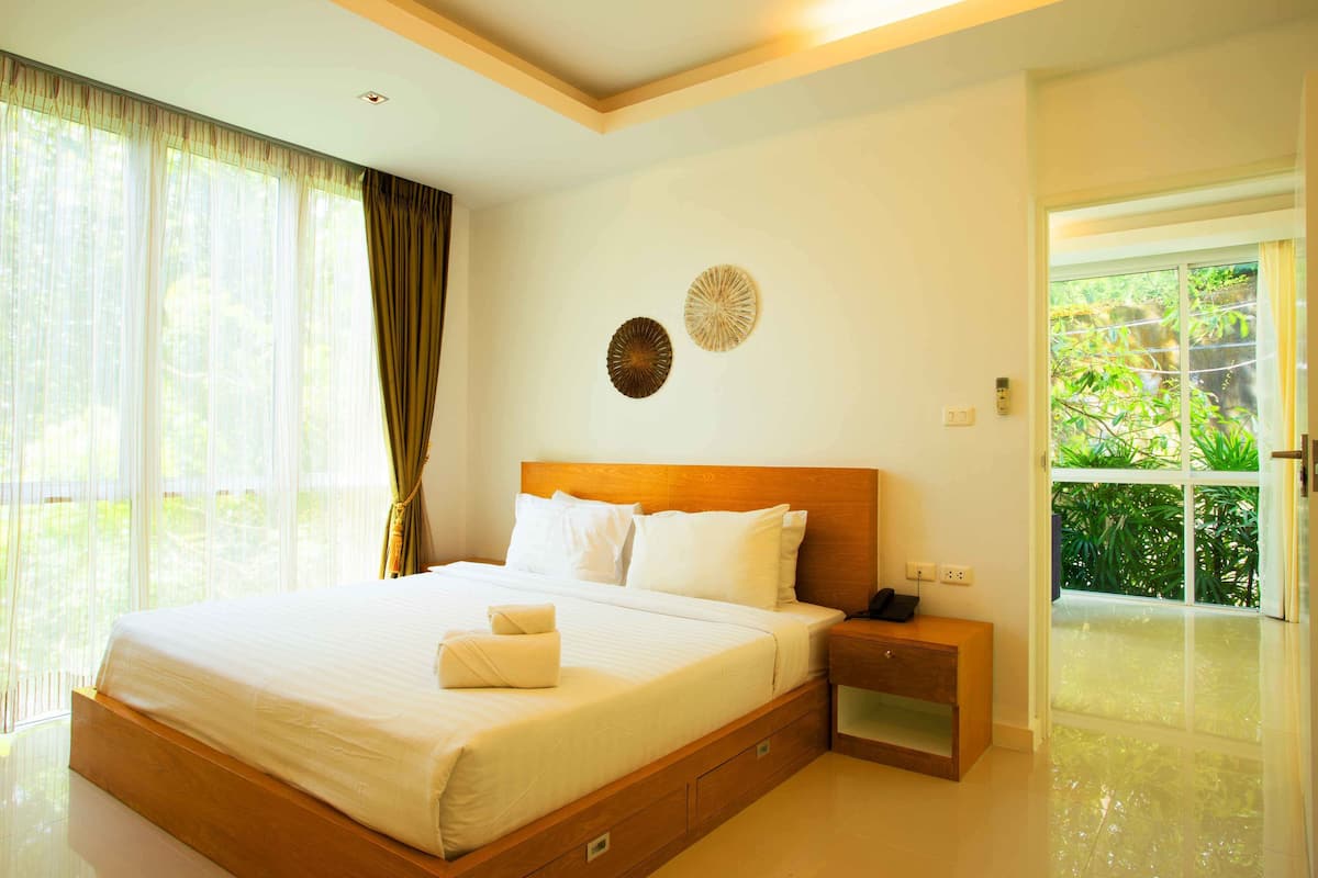 Quarto de hotel com cama de madeira, roupa de cama branca, mesa de cabeceira com abajur e vista para o verde pela janela.