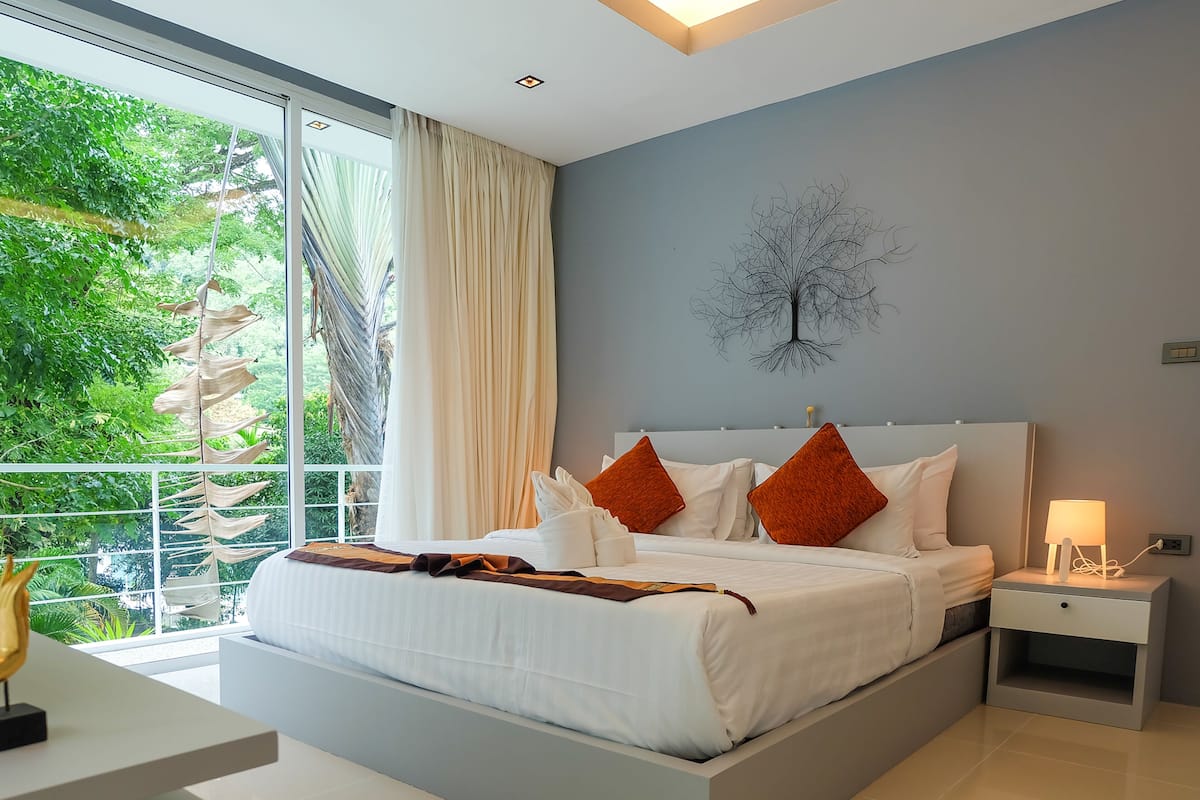 Quarto moderno com uma cama grande, mesa de cabeceira e vista para o verde através de uma janela ampla.