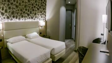 Quarto de hotel com duas camas, uma escrivaninha, uma cadeira, televisão e porta que dá para outro espaço.