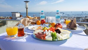Mesa com itens de café da manhã, incluindo frutas, queijo e bebidas, com vista para o mar.