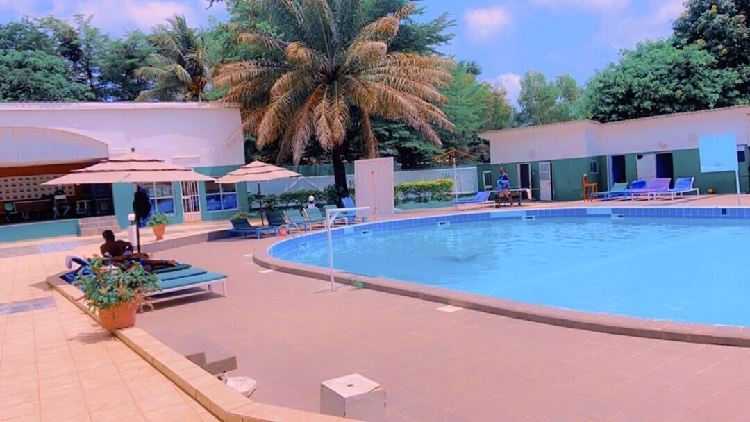 Piscina com espreguiçadeiras e guarda-sóis, rodeada por um edifício com telhado curvo e palmeiras.