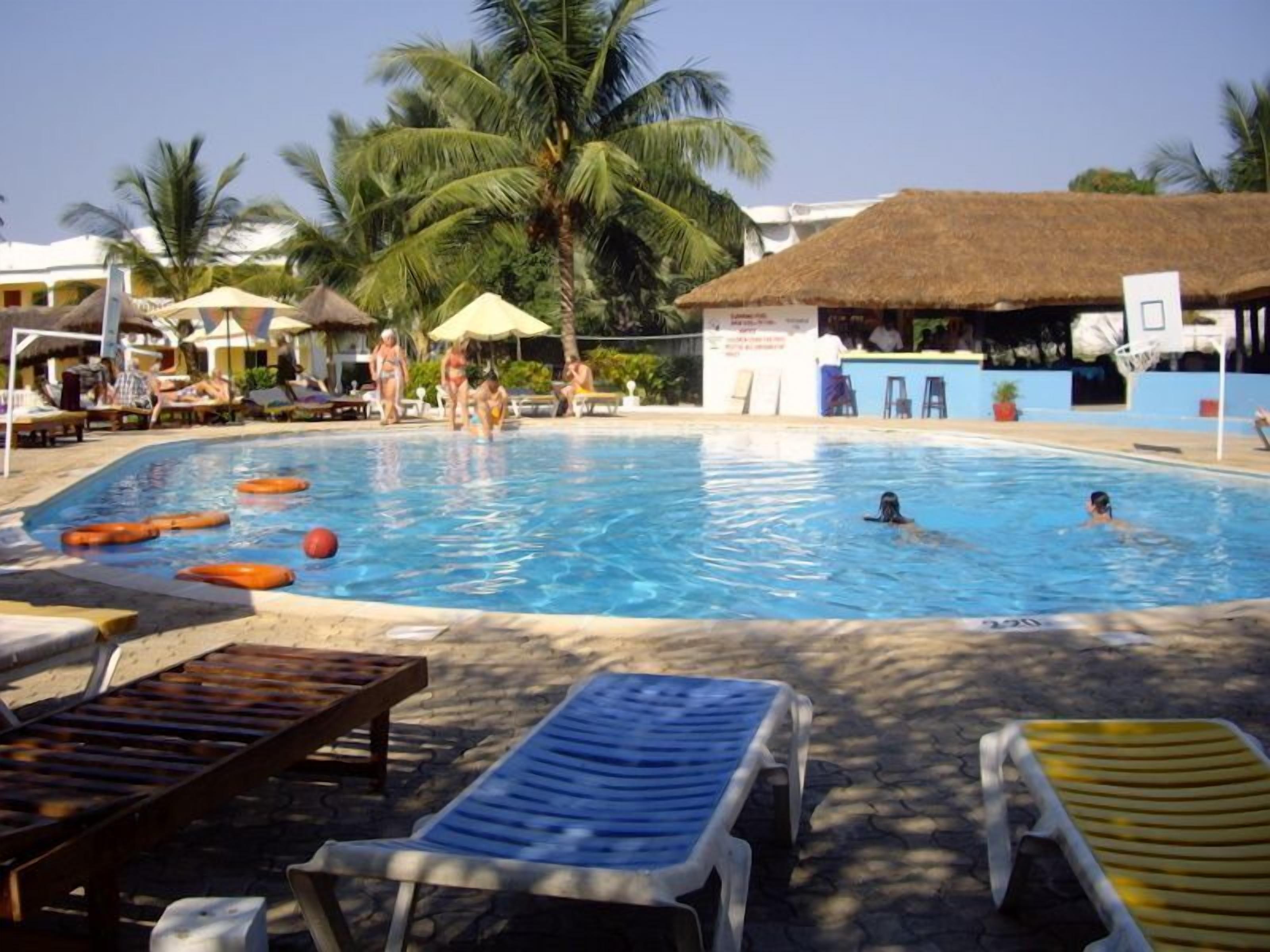 Piscina com espreguiçadeiras, palmeiras e um edifício com telhado de palha.