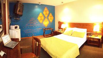 Quarto de hotel com cama, escrivaninha com laptop, cadeira, mesa de cabeceira com abajur e parede com texto e gráficos.