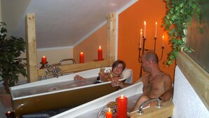 Um quarto de spa com duas banheiras separadas, cada uma com estrutura de madeira e um espelho acima. Há velas no chão e na parede, e uma planta pendurada na parede.