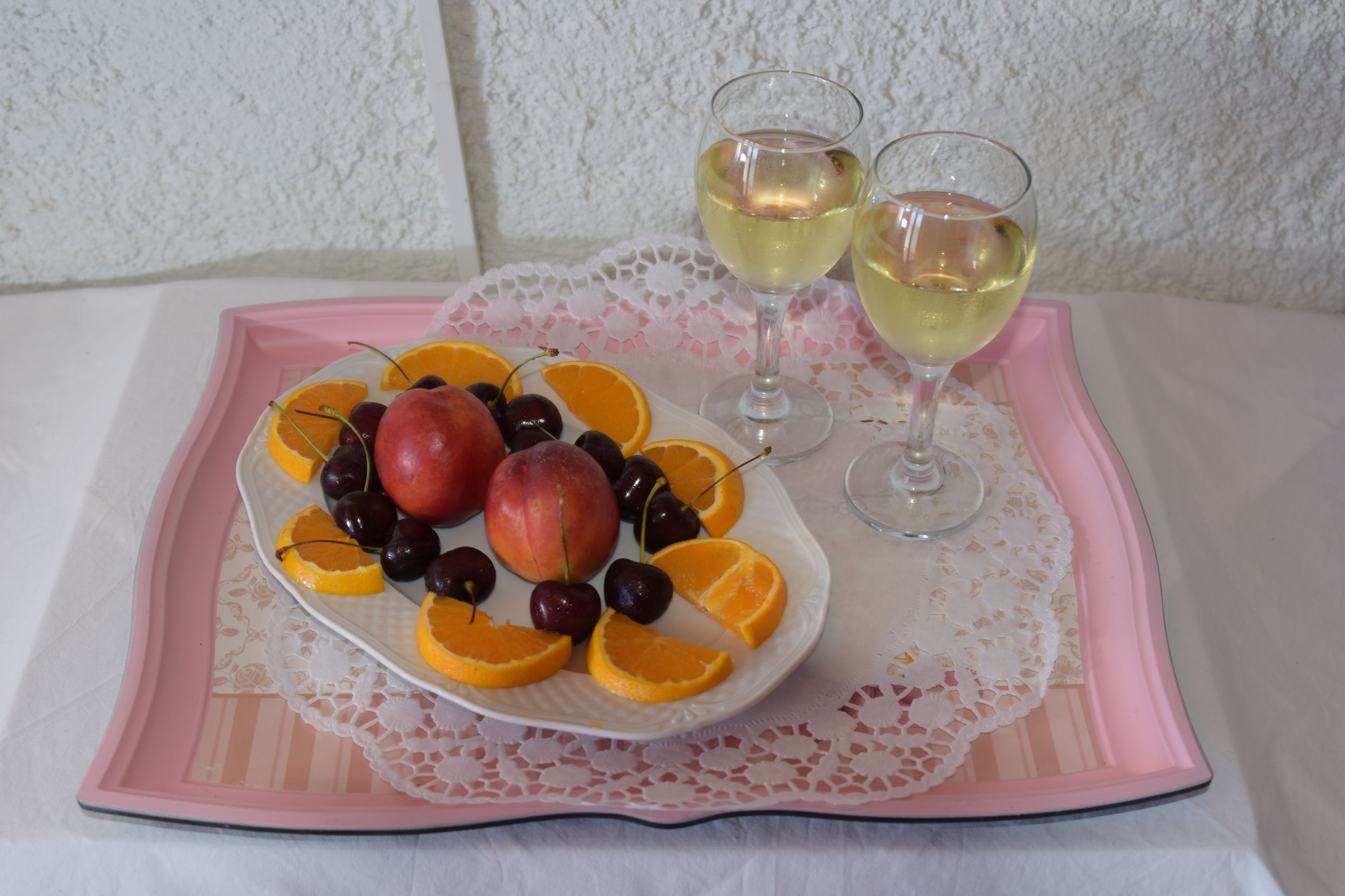 Uma tábua de frutas com fatias de laranja, maçã e cerejas em um prato rosa, acompanhada por dois copos de vinho branco sobre uma toalha de renda.