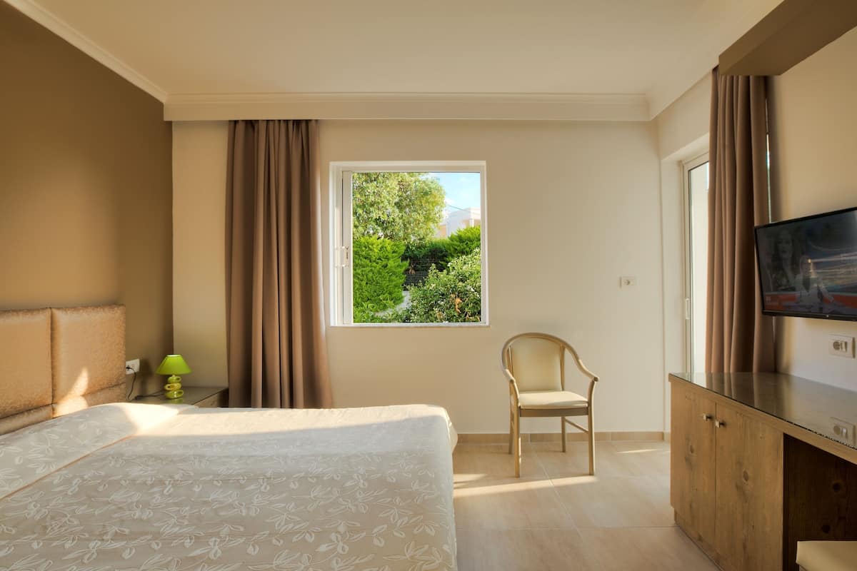 Quarto de hotel com cama, cadeira, televisão e janela com vista para o verde.