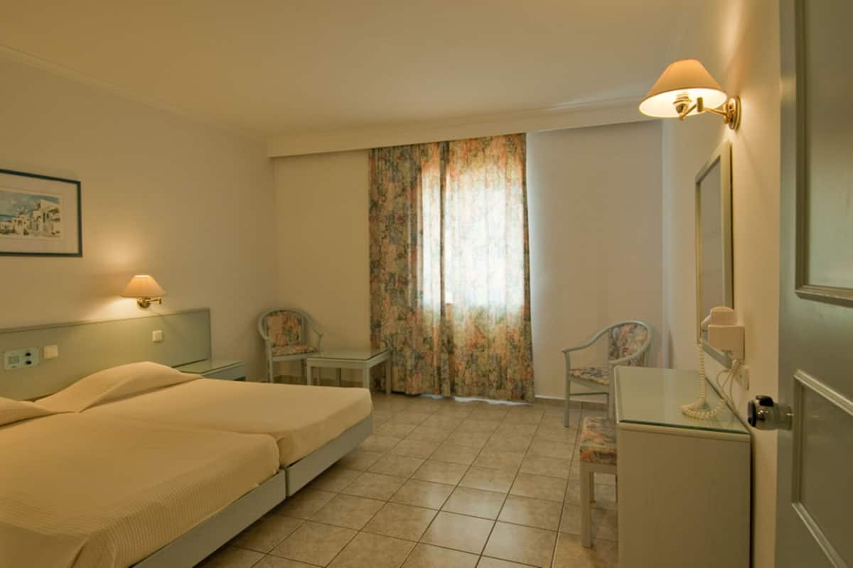 Quarto de hotel com duas camas, uma escrivaninha, uma cadeira, um telefone e uma janela com cortinas.