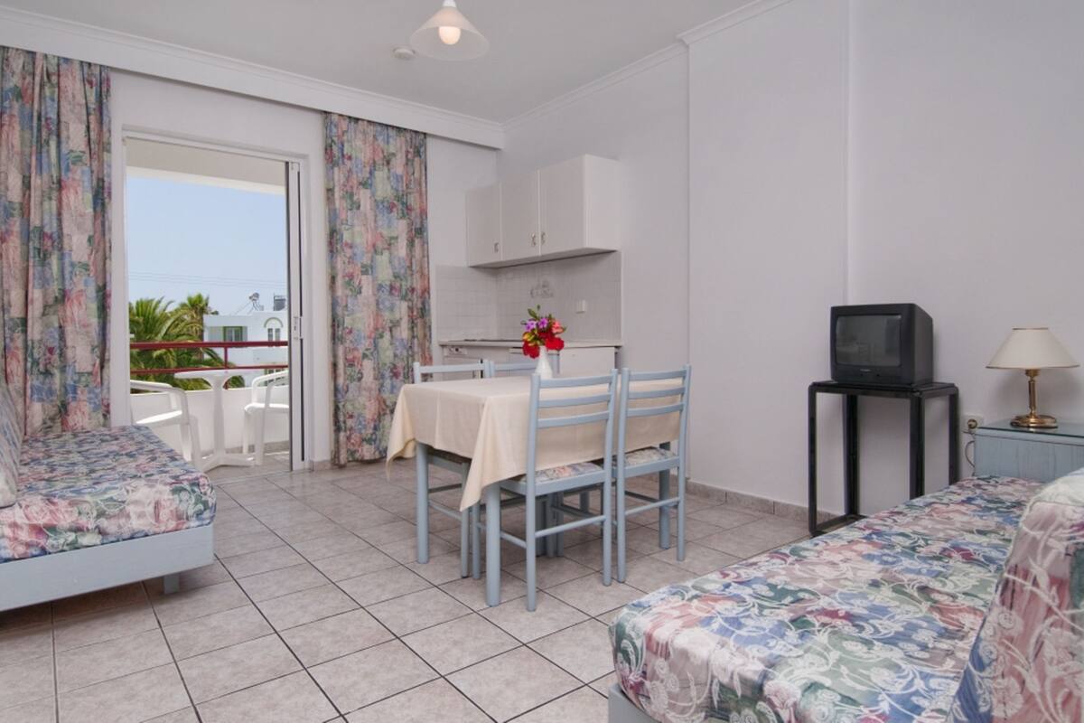 Quarto com cama, mesa com cadeiras, televisão e varanda com mesa e cadeiras.