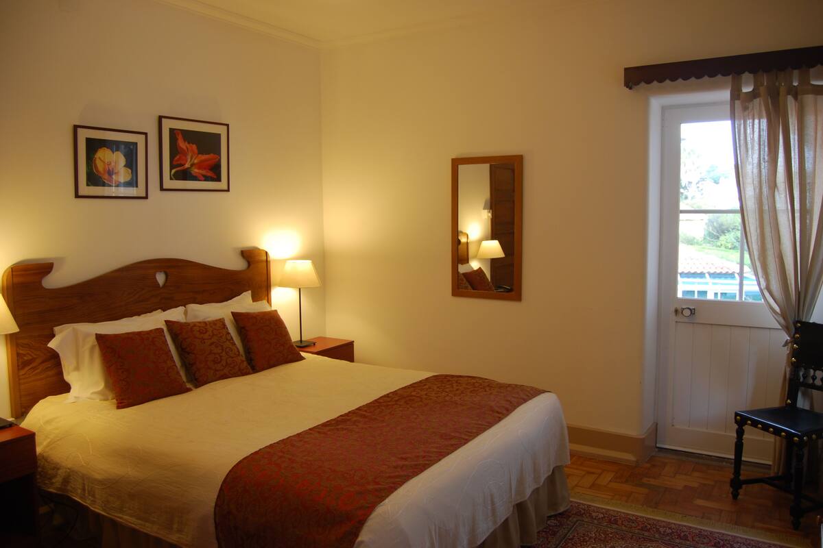 Quarto de hotel com uma cama grande, duas luminárias de cabeceira, um espelho e vista para o exterior através de uma porta.