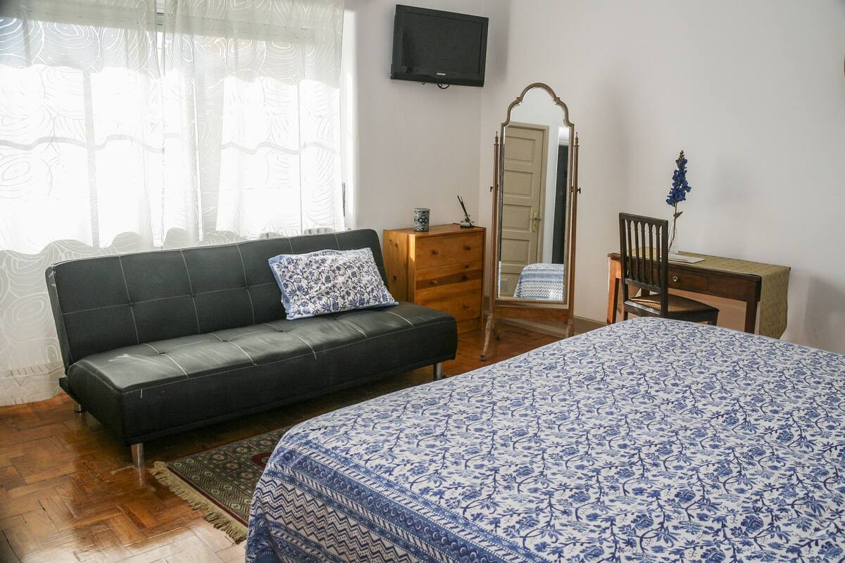 Quarto com cama, sofá, cômoda, espelho e televisão na parede.