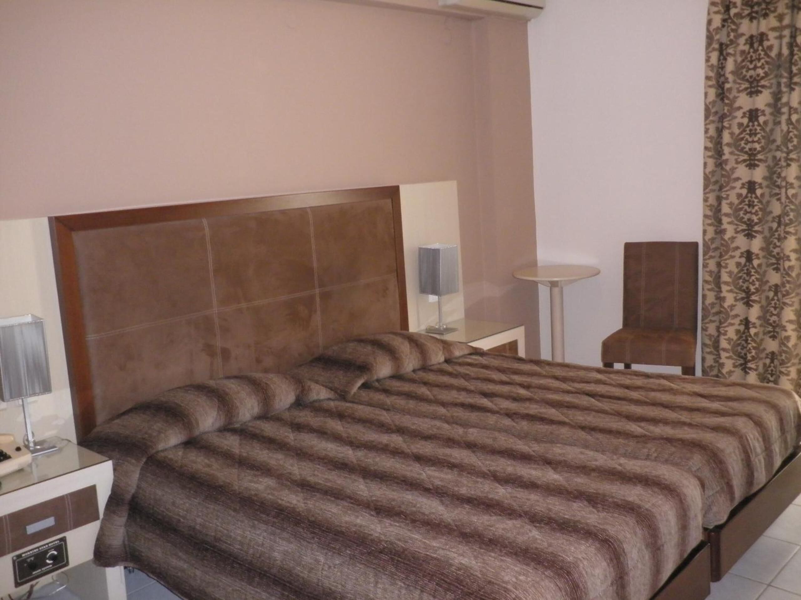 Quarto com cama grande, mesa de cabeceira, luminária, cadeira e pequena mesa.