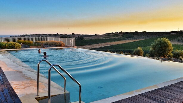 Uma piscina infinita com deck de madeira e grades de metal, rodeada por colinas ajardinadas e um céu claro.