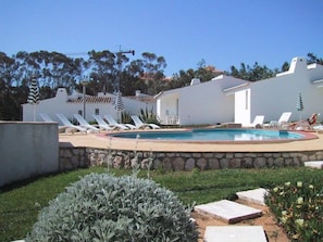 Área de piscina com espreguiçadeiras, parede de pedra e um edifício branco ao fundo.