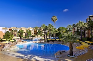 Área de piscina em resort, com espreguiçadeiras e guarda-sóis, rodeada por palmeiras e edifícios do resort.