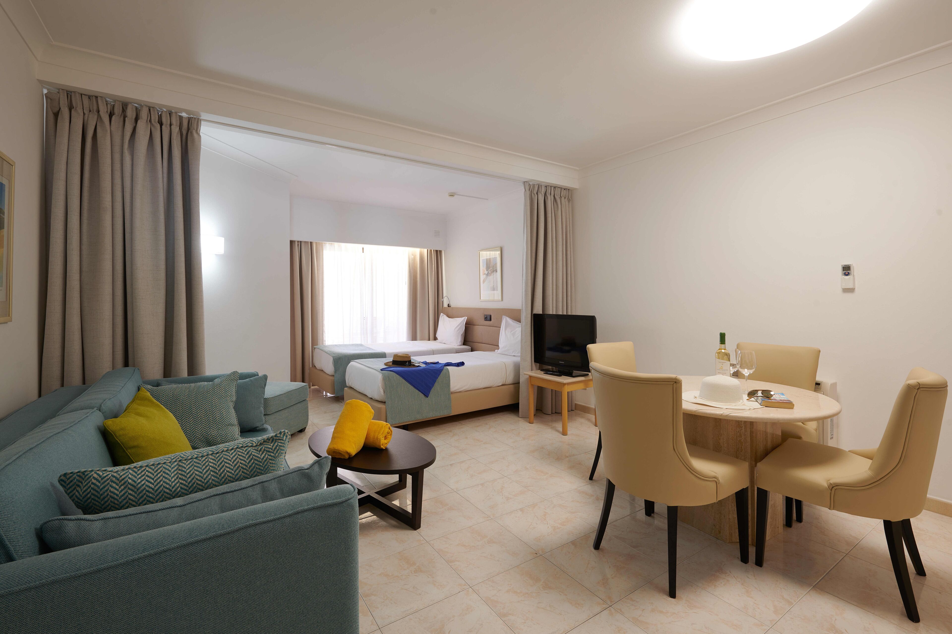 Quarto de hotel com sofá, cama, televisão, mesa de jantar com cadeiras e uma pequena mesa lateral.