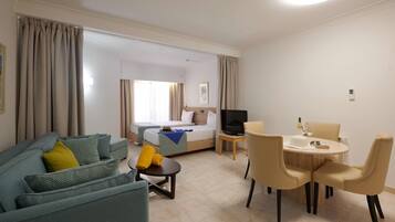 Quarto de hotel com sofá, cama, televisão, mesa de jantar com cadeiras e uma pequena mesa lateral.