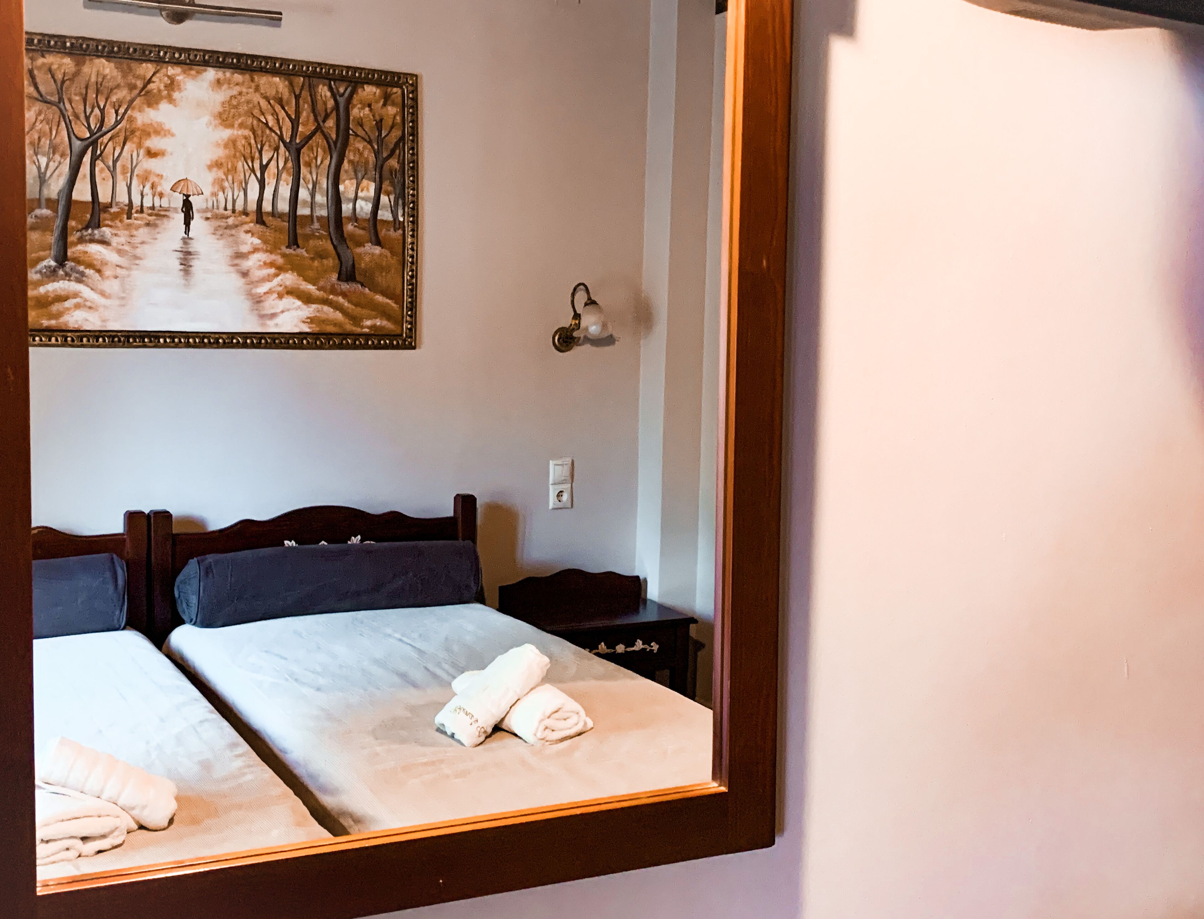 Quarto de hotel com duas camas, um espelho grande, um quadro de uma trilha arborizada e uma mesa de cabeceira com abajur.