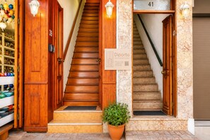 Entrada de hotel com escadaria de madeira, placa anunciando quartos para alugar e um vaso de planta.