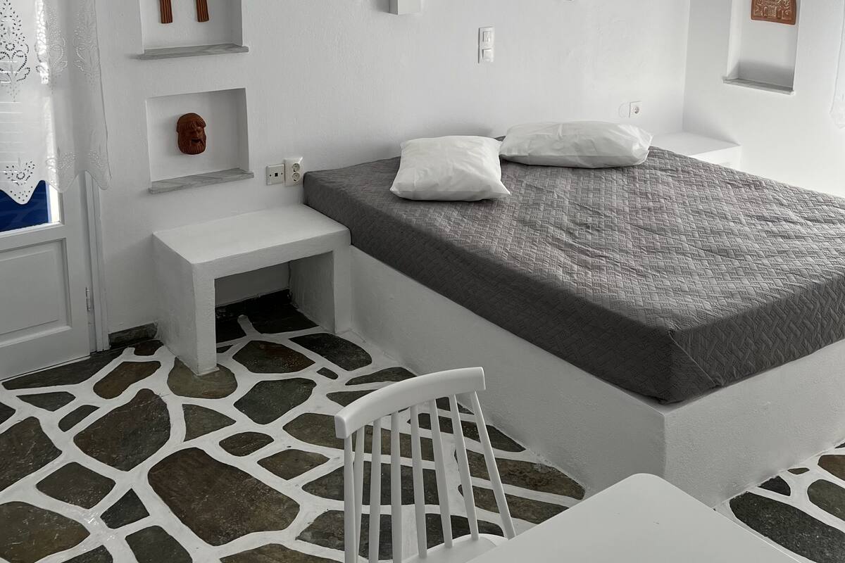 Quarto minimalista com uma cama, uma cadeira branca, uma mesinha e objetos decorativos nas prateleiras.