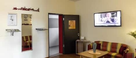 Uma sala de estar com piso de madeira, um tapete vermelho, um sofá, uma televisão e uma porta que dá para outro cômodo.