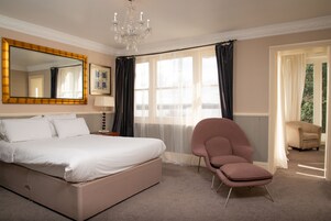 Quarto de hotel com uma cama grande, uma poltrona, um candelabro e uma janela com cortinas.