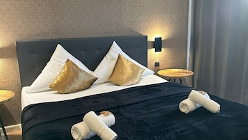 Cama bem arrumada com colcha preta, duas almofadas douradas e duas toalhas brancas.
