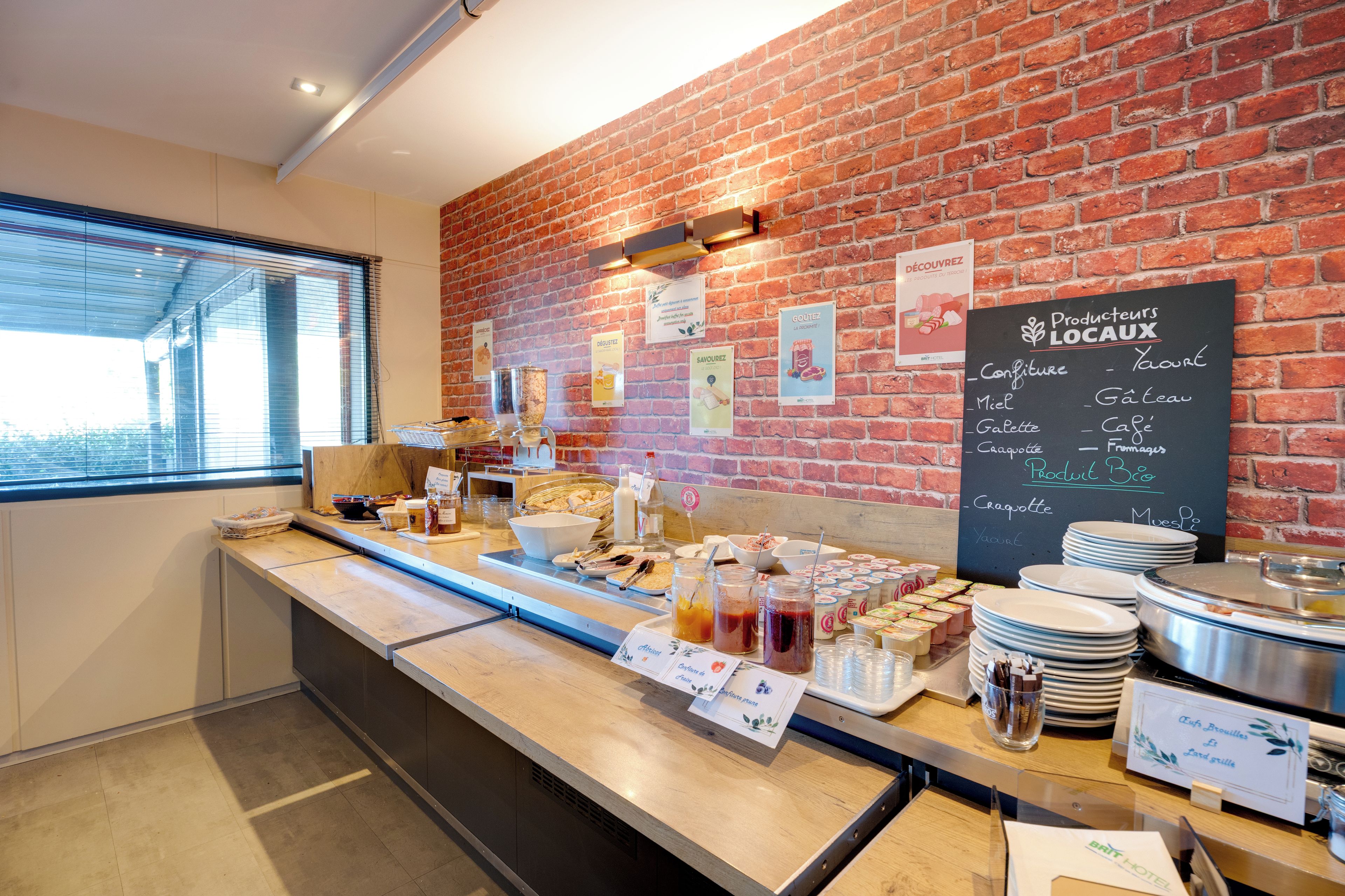 Espaço de buffet de café da manhã com parede de tijolos, diversos alimentos e cardápio em lousa.
