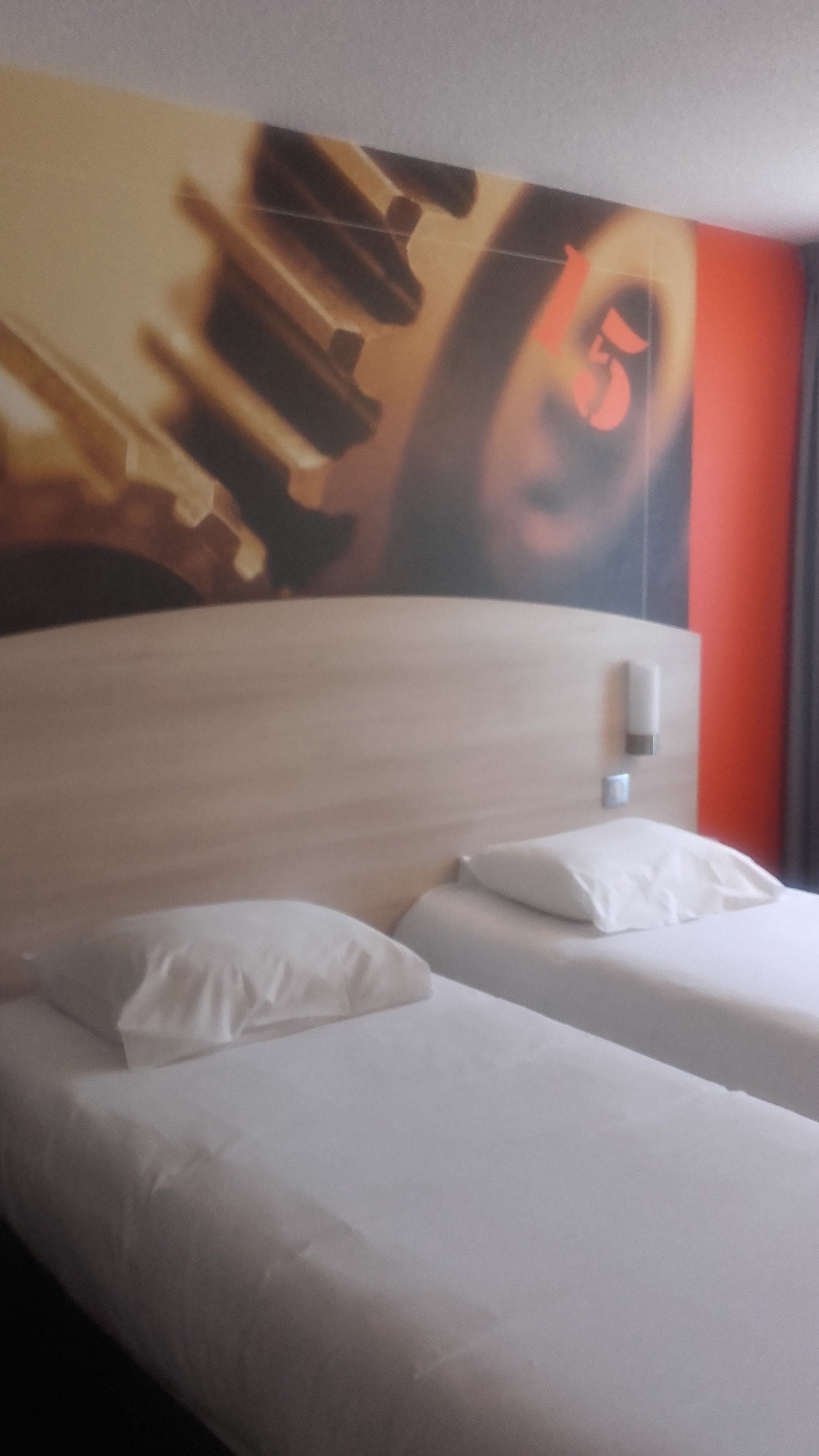 Quarto de hotel com duas camas de solteiro, cabeceira de madeira, luz noturna fixada na parede e vista parcial de uma cortina vermelha.