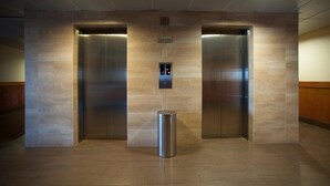 Uma área de elevadores moderna, com dois elevadores, uma lixeira e um aviso para não usar o vaso sanitário.