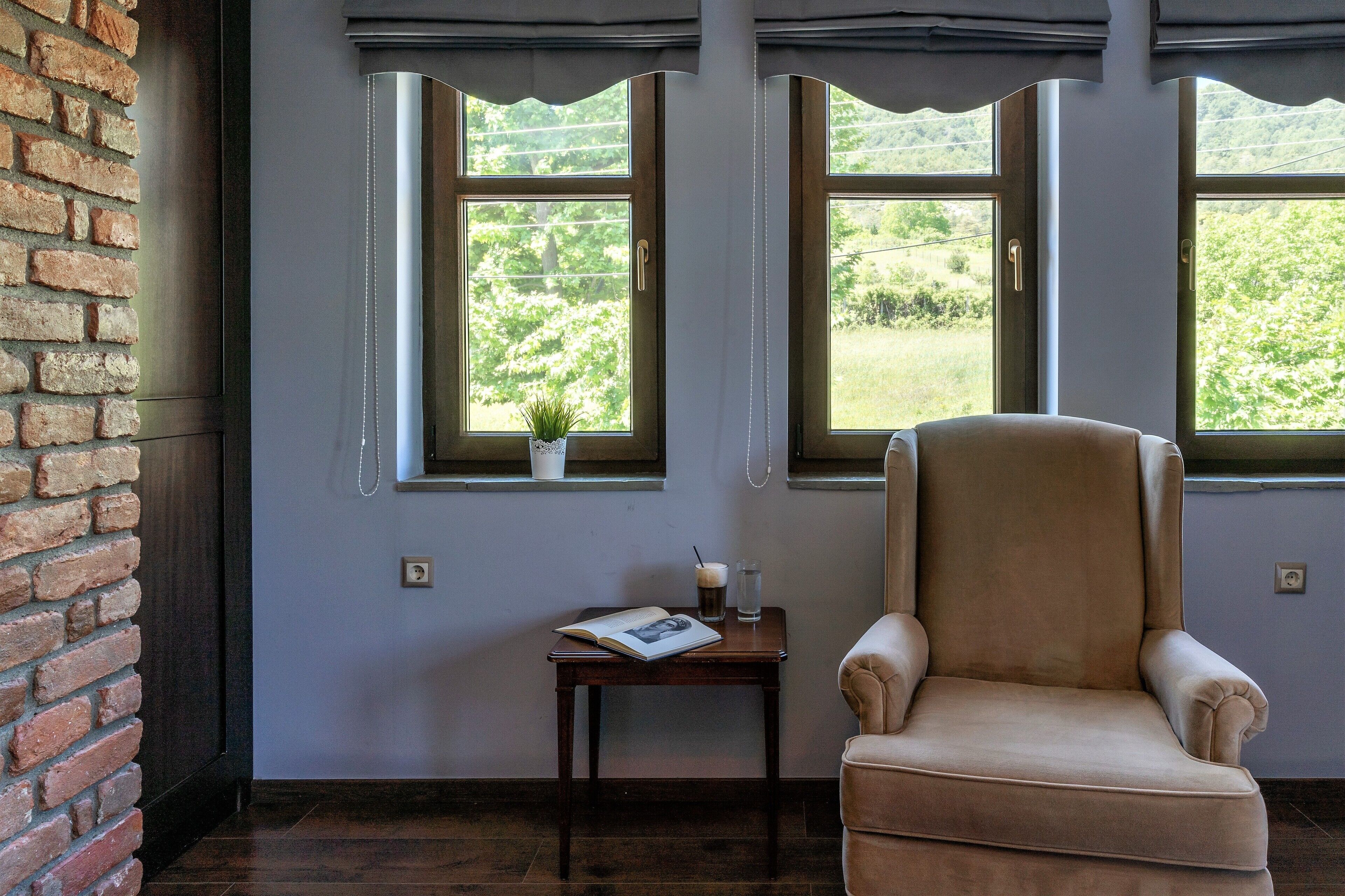 Quarto com parede de tijolos, duas janelas com cortinas, uma mesinha com um livro e um copo, e uma poltrona bege.