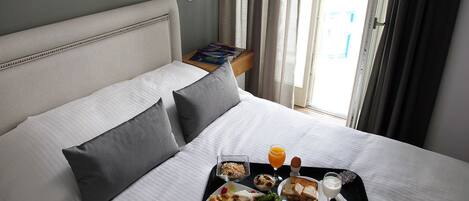 Uma cama bem arrumada com uma bandeja de café da manhã, incluindo ovos, torradas e suco, em um quarto com vista para a cidade através de uma janela.