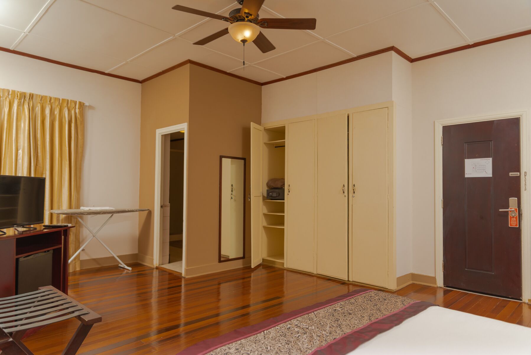 Quarto com piso de madeira, ventoinha no teto, televisão, tábua de passar, guarda-roupa e uma porta.