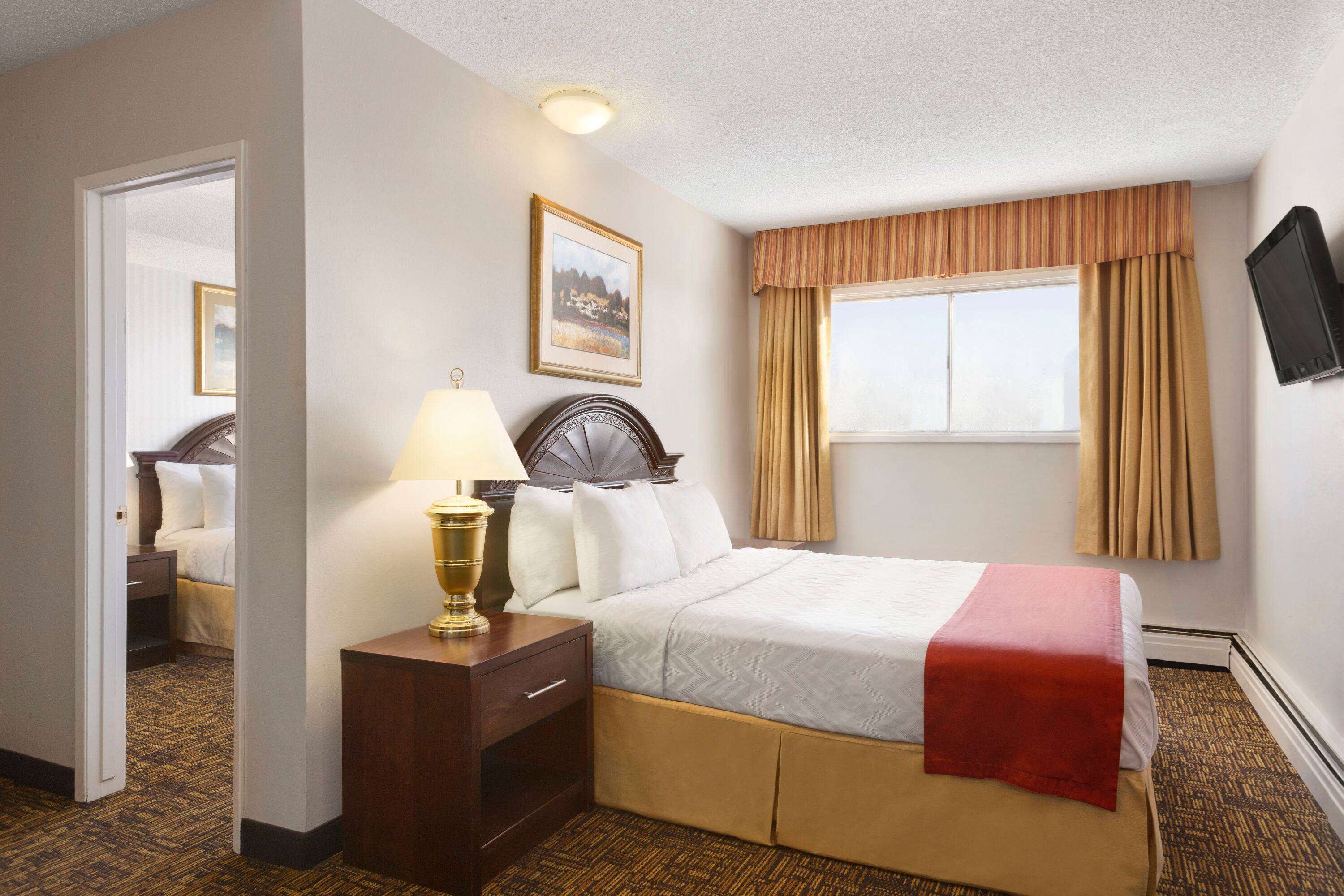 Quarto de hotel com cama, mesa de cabeceira com abajur, quadro na parede e janela com cortinas.