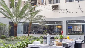 Espaço de refeições ao ar livre com mesas preparadas para o café da manhã, incluindo cafeteiras, pratos e uma cesta de frutas.