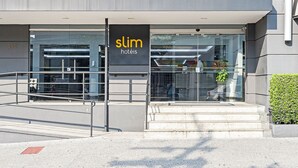 Entrada moderna de hotel com fachada de vidro, uma placa com o texto "Slim Hotels" e uma escadaria que leva à entrada.