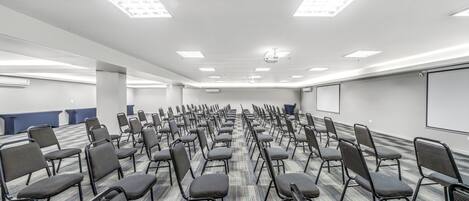 Uma sala de conferências com fileiras de cadeiras, uma tela de projetor e piso coberto de carpete.
